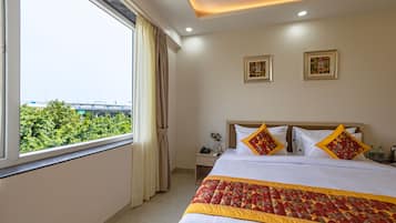 Quarto de hotel com uma cama grande, duas obras de arte emolduradas na parede, uma mesa de cabeceira com telefone e um vaso, e uma janela com cortinas.