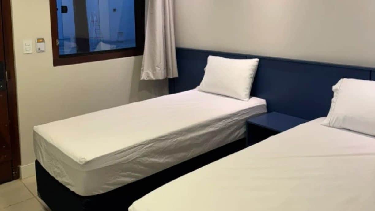Quarto de hotel com duas camas de solteiro, uma pequena mesa de cabeceira, uma janela com cortinas e uma porta.