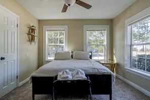 Quarto com cama, duas janelas, ventilador de teto e uma pequena mesa com uma planta.
