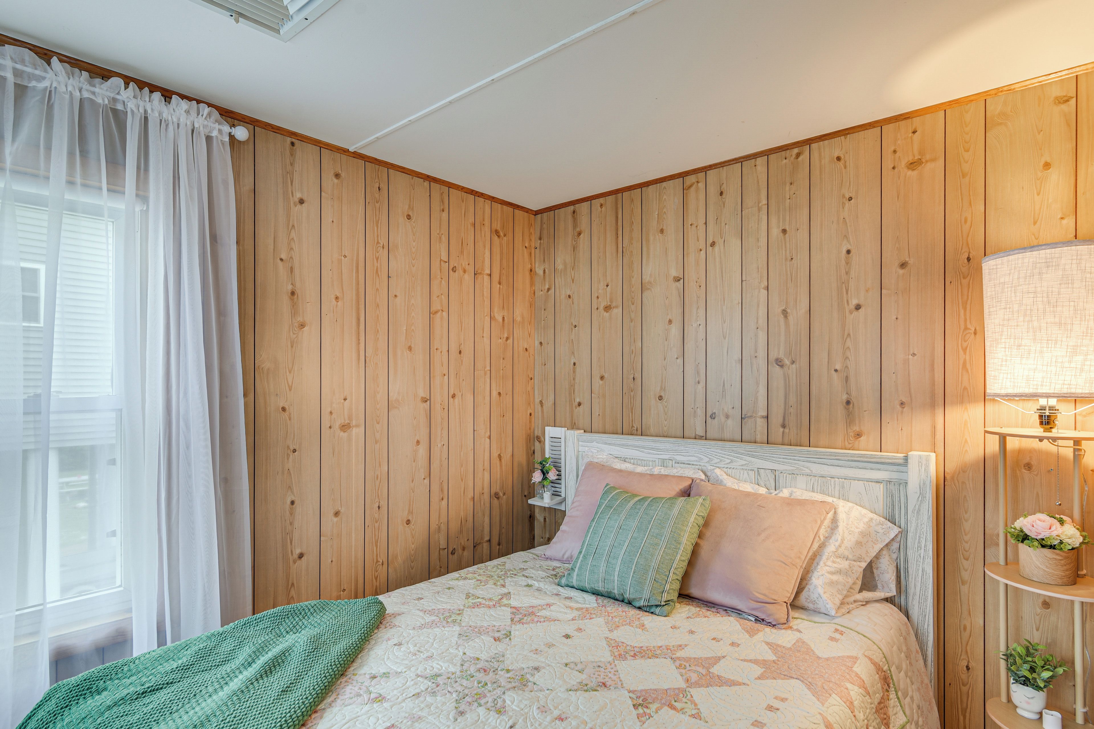 Quarto com paredes de madeira, cama com roupa de cama floral, mesa de cabeceira com abajur e janela com cortinas leves.