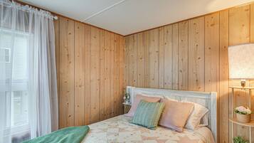 Quarto com paredes de madeira, cama com roupa de cama floral, mesa de cabeceira com abajur e janela com cortinas leves.