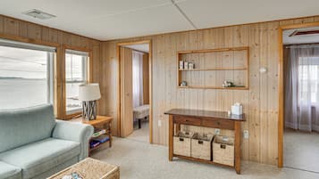 Quarto com parede revestida de madeira, um sofá, uma escrivaninha com cestos organizadores e vista para o mar pela janela.