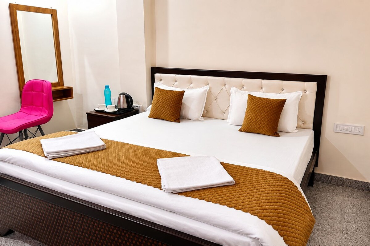 Quarto de hotel com cama, cadeira, espelho e mesa de cabeceira com chaleira.