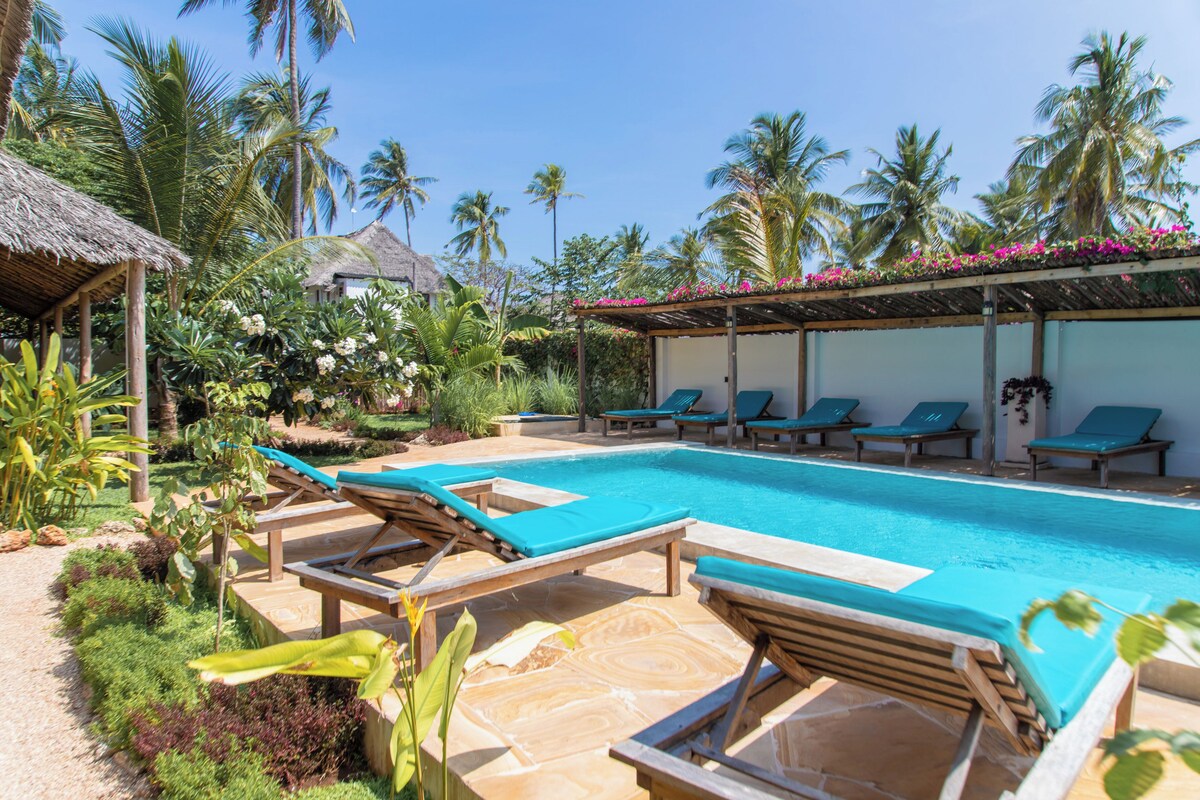 Área de piscina com espreguiçadeiras e uma estrutura com telhado de palha.