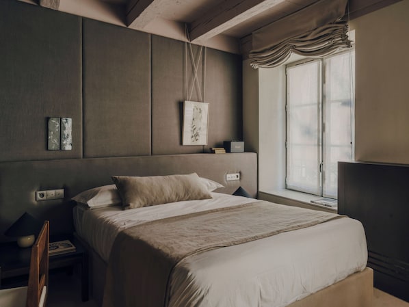 Quarto com uma cama grande, mesa de cabeceira, abajur, janela com cortinas e uma obra de arte na parede.