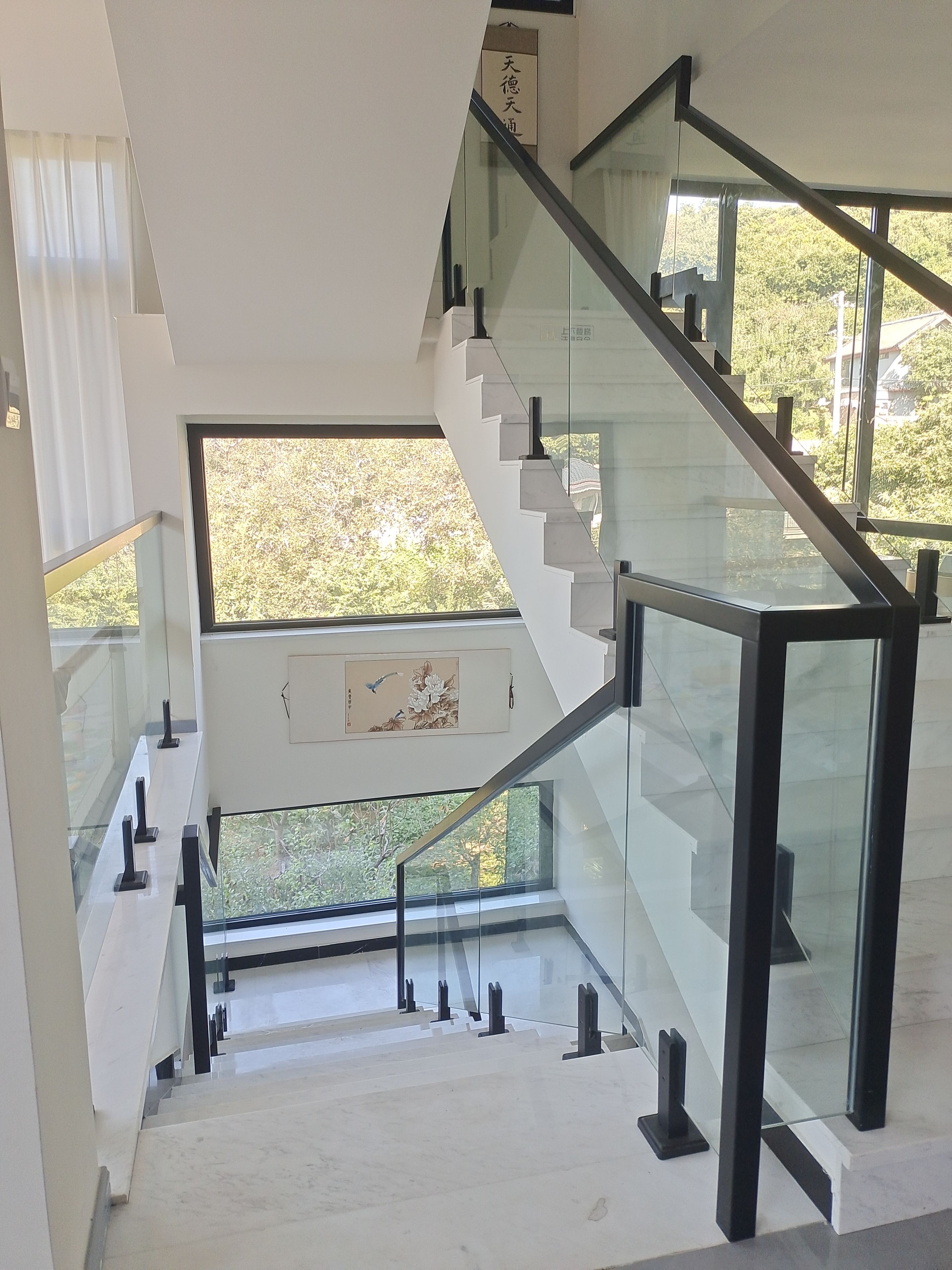 Uma escadaria moderna com guarda-corpos de vidro e vista para o verde lá fora.