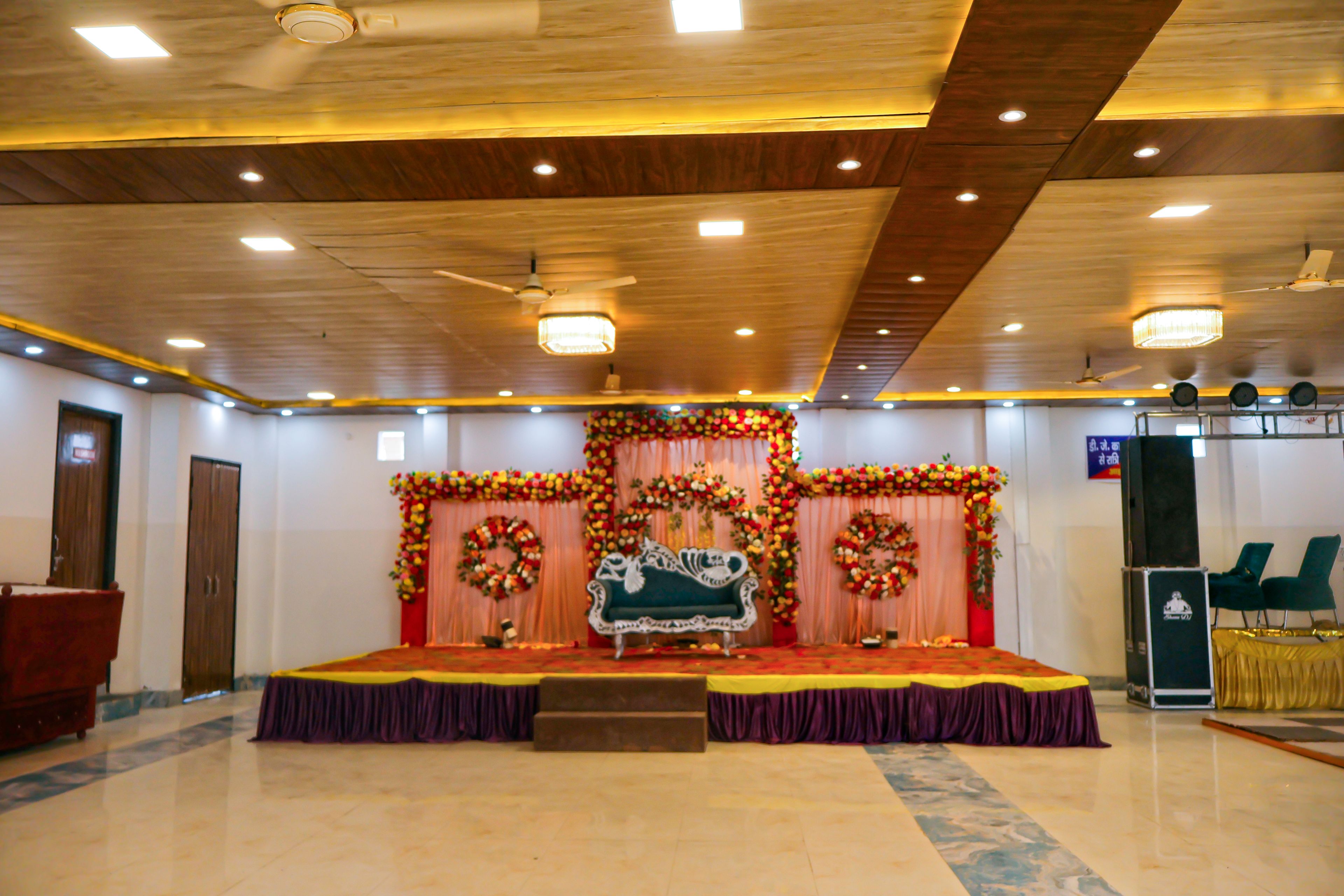 Palco com decorações florais e um sofá, teto com painéis de madeira e iluminação, e área de estar com cadeiras.