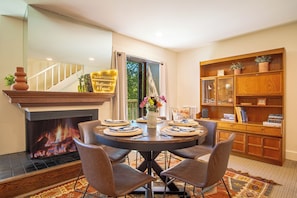 Uma sala de jantar com lareira, uma mesa redonda posta para quatro pessoas e uma estante com livros e objetos decorativos.