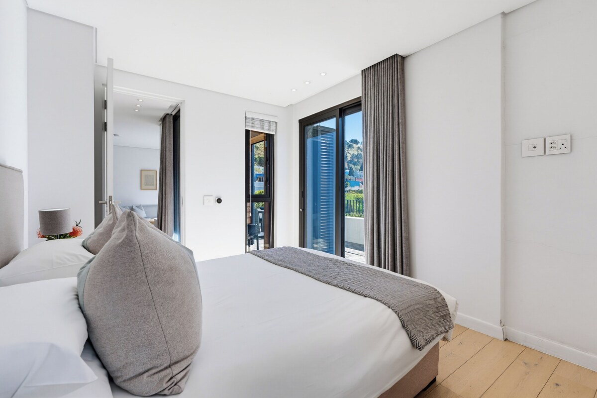 Quarto moderno com uma cama grande, mesa de cabeceira e vista para o exterior através de uma porta de vidro.