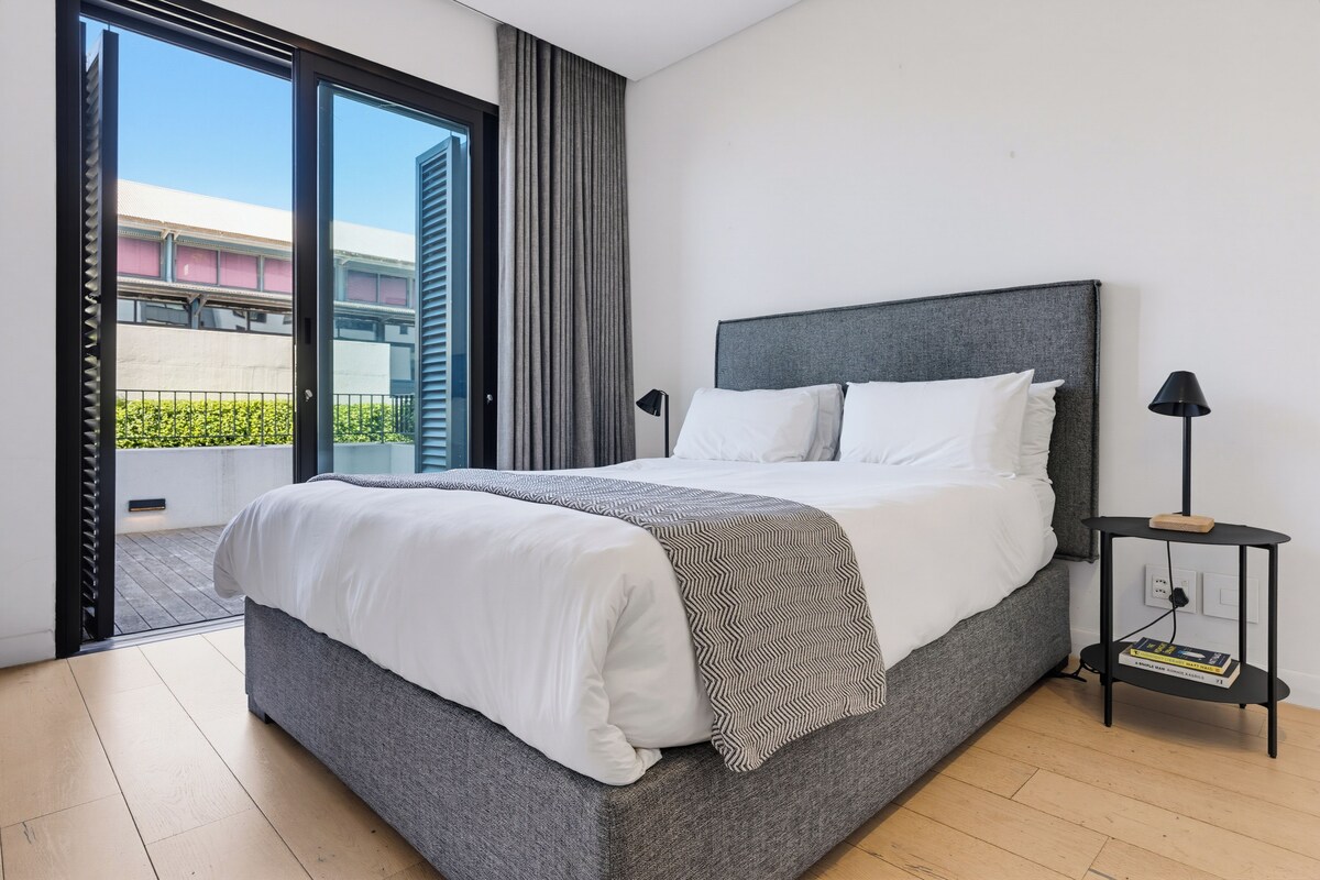 Quarto moderno com uma cama grande, uma mesa de cabeceira, um abajur e vista para área externa por uma porta de correr.