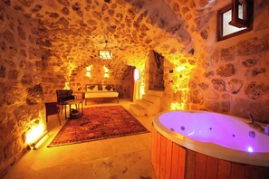 Quarto com paredes de pedra, uma banheira de madeira, uma cama e uma pequena mesa com cadeiras.