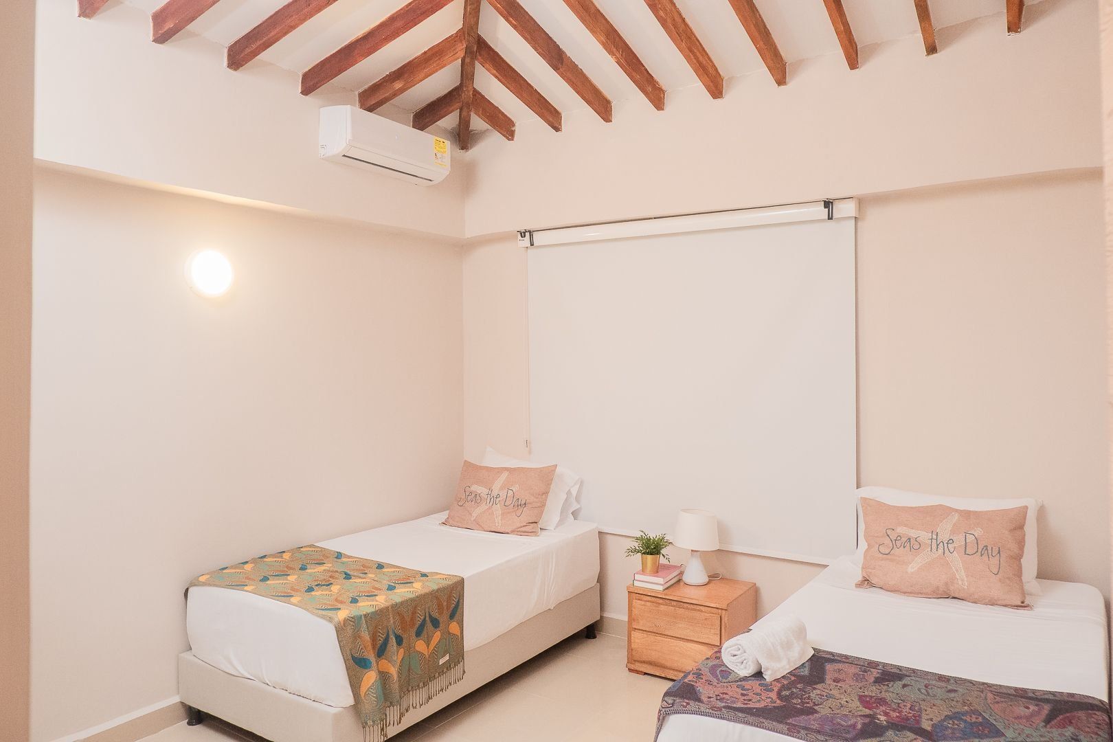 Quarto com duas camas de solteiro, mesa de cabeceira com luminária, pequena mesa com livros e planta, e ar-condicionado na parede.
