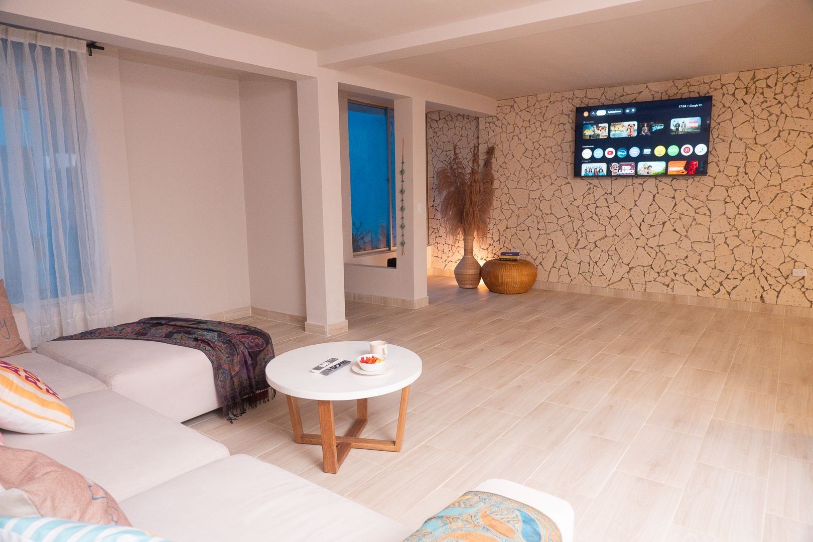 Uma sala de estar moderna com um sofá, uma mesa redonda pequena com um controle remoto e uma TV de tela plana fixada em uma parede texturizada.