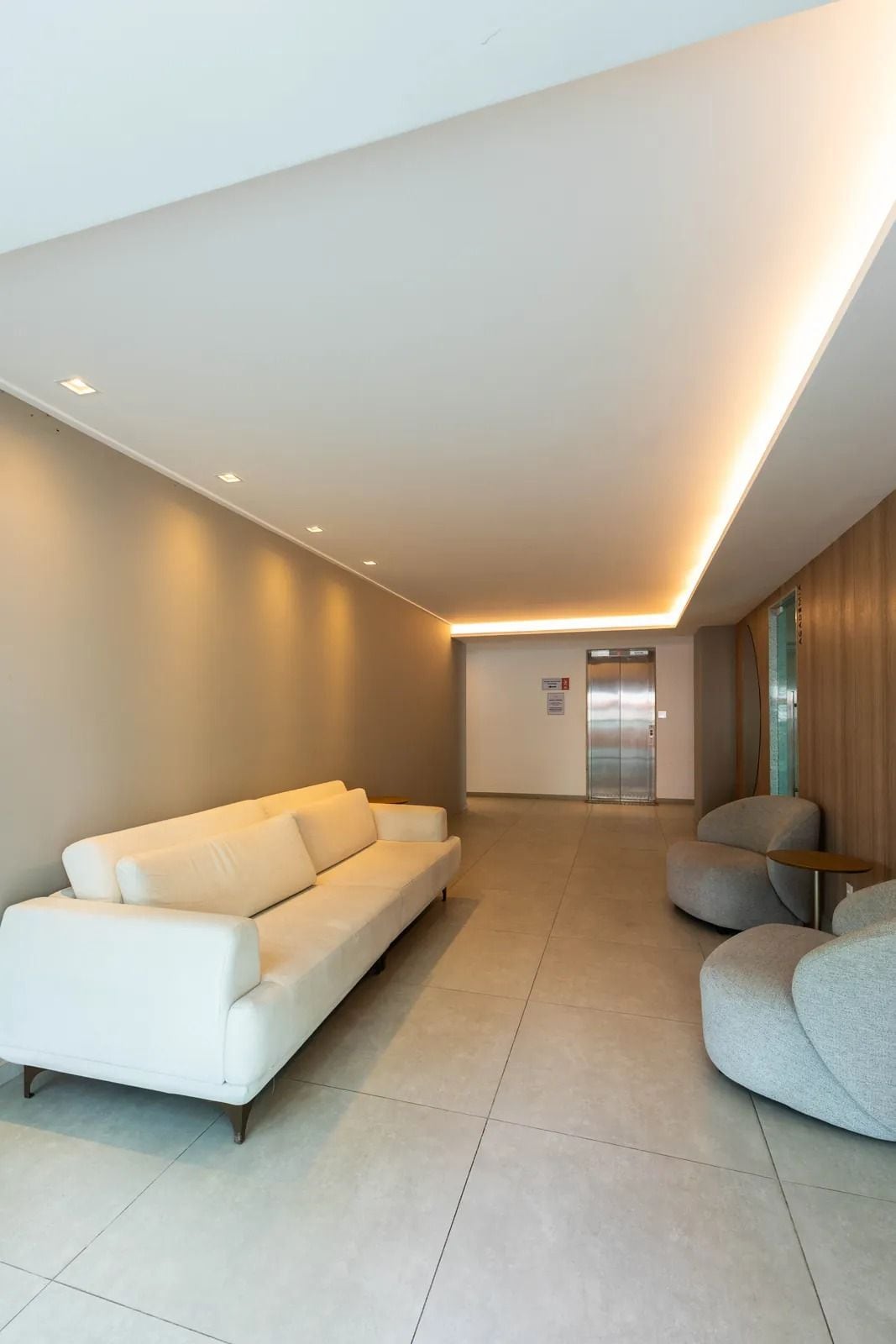 Uma área de espera moderna com um sofá branco, uma poltrona cinza e uma mesa pequena.
