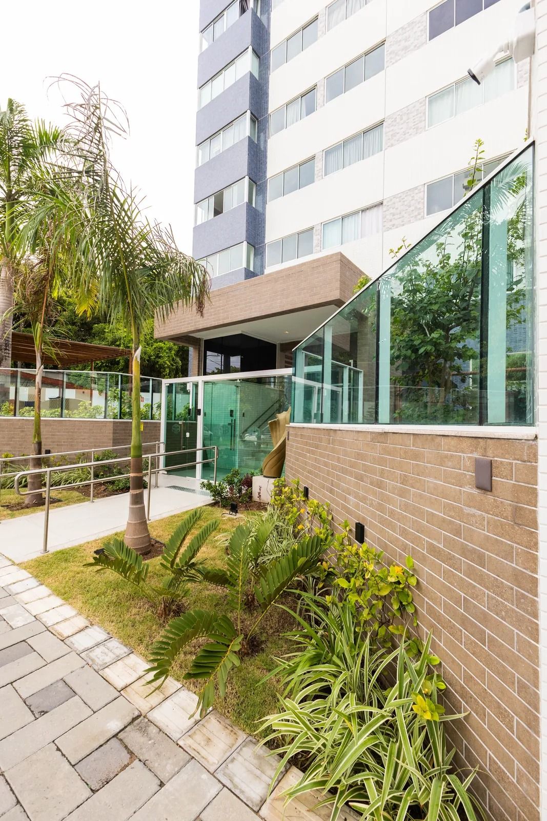 Um edifício moderno com fachada de vidro, exterior em tijolo e área ajardinada com palmeiras e plantas.