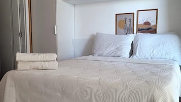 Quarto com cama, roupa de cama branca, um guarda-roupa e duas imagens emolduradas na parede.