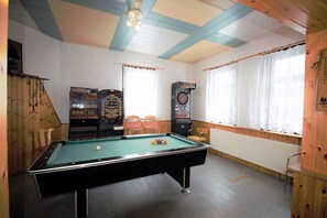Uma mesa de bilhar em um ambiente com paredes de madeira e teto com vigas azuis. Há duas jukeboxes e um alvo de dardos no espaço.