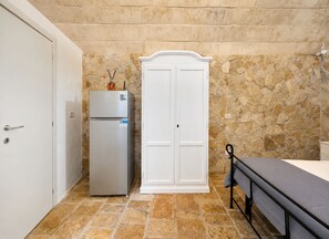 Quarto compacto com guarda-roupa branco, um frigorífico pequeno e cama com estrutura de metal.