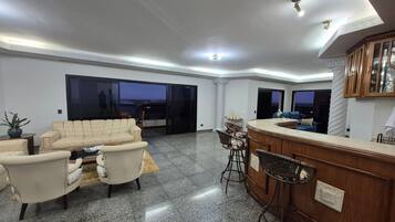 Ampla sala de estar com sofá, cadeiras, espaço bar e vista através de grandes janelas.