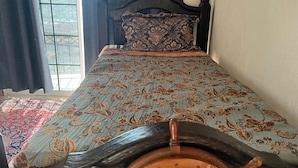 Cama com colcha estampada e travesseiro decorativo, cabeceira de madeira e vista pela janela.