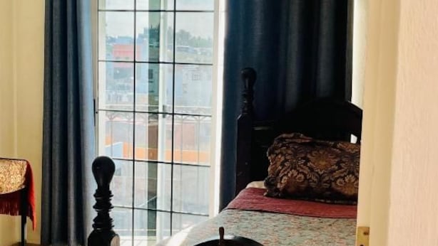 Quarto com cama e cabeceira em formato de volante, janela com cortinas e vista para prédios.