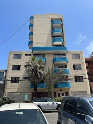 Um edifício de vários andares com varandas, uma palmeira na frente e carros estacionados.