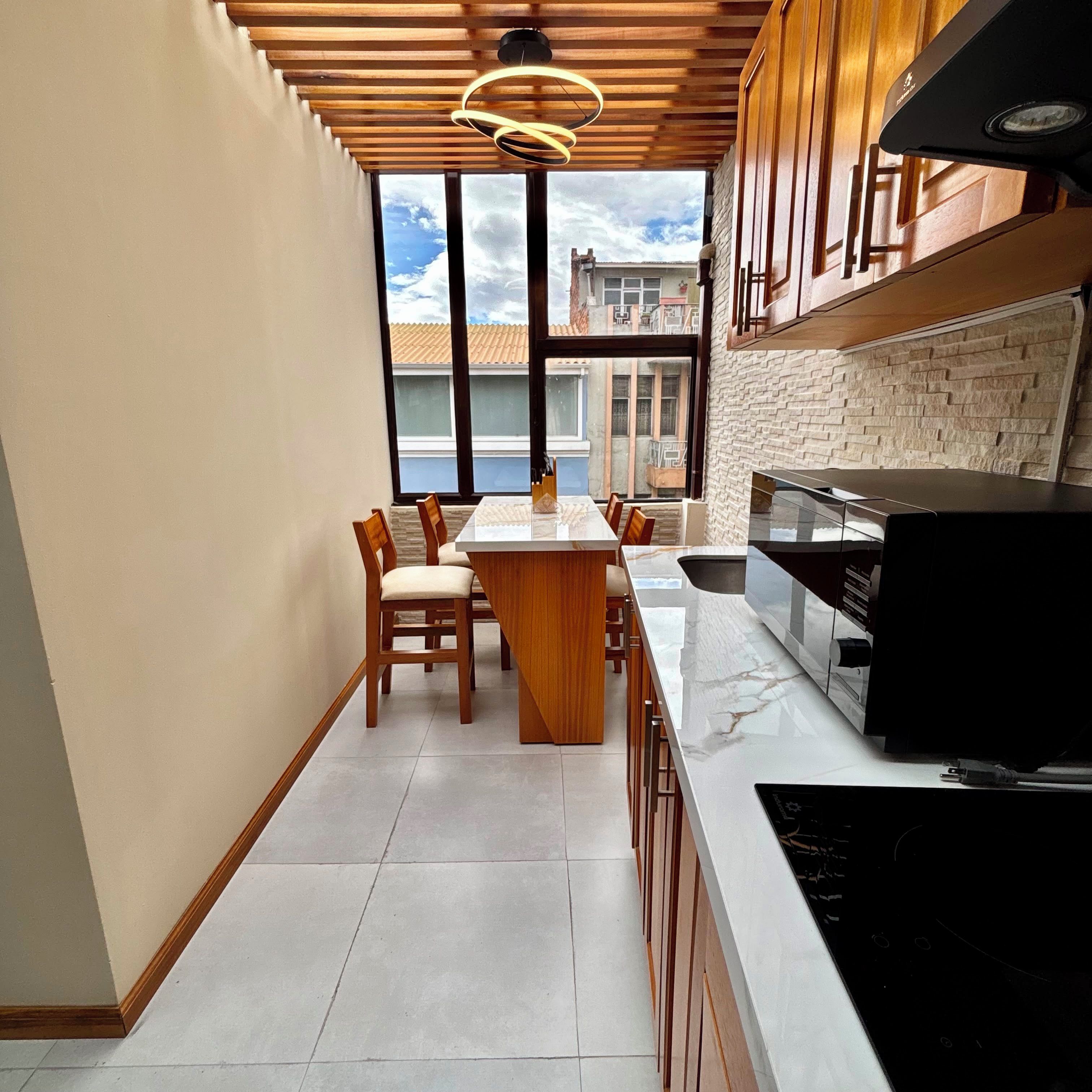 Cozinha moderna com ilha central, cadeiras de madeira, forno embutido e uma grande janela com vista para o exterior.