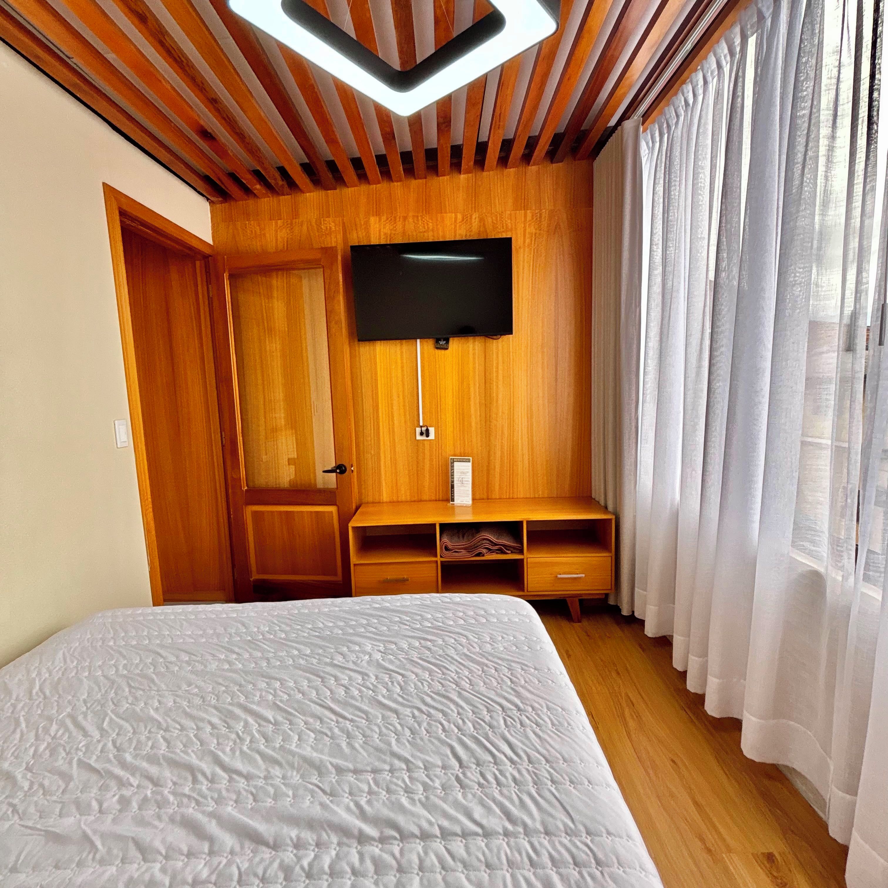 Quarto com cama, televisão na parede, guarda-roupa de madeira e janela com cortinas.