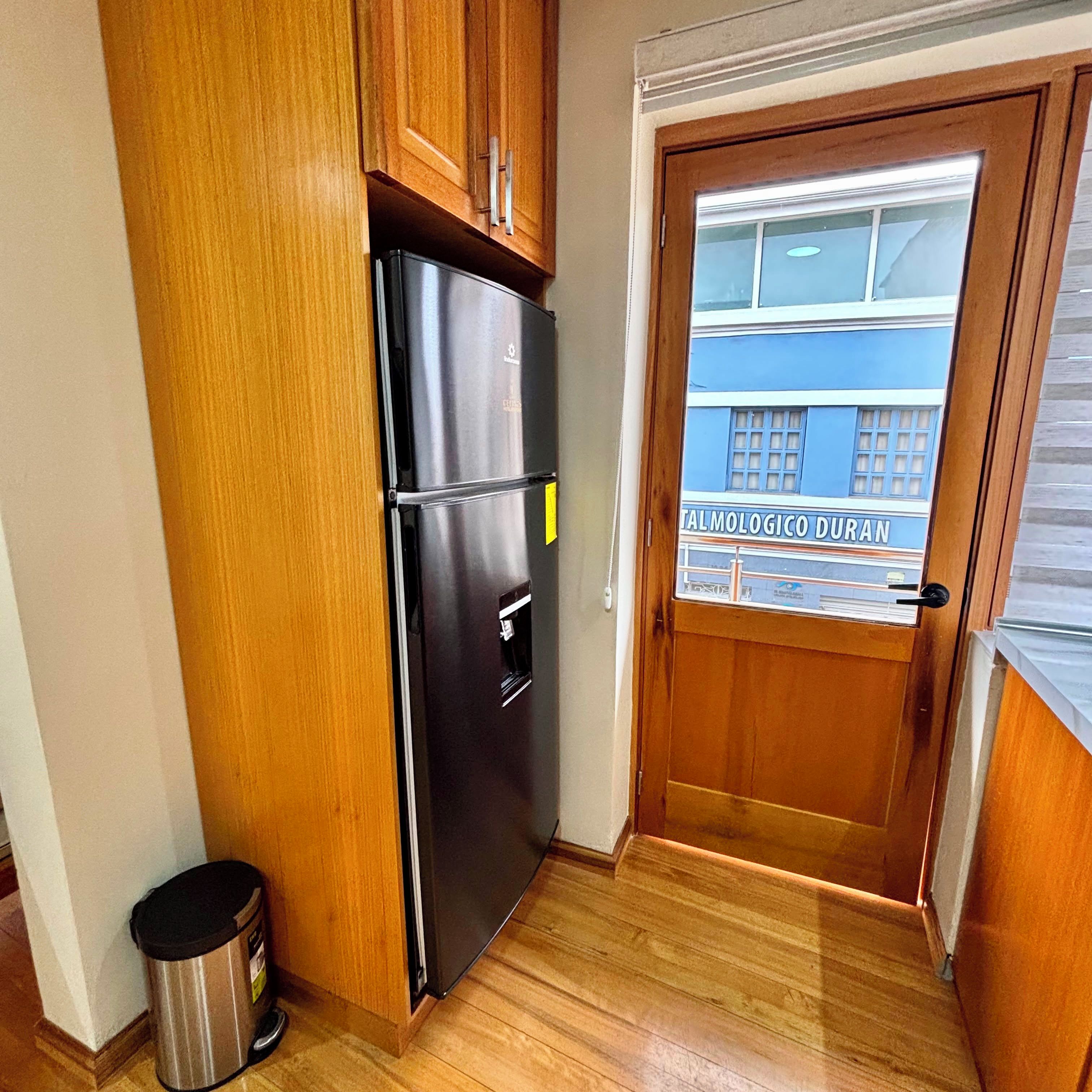 Cozinha compacta com armários de madeira, geladeira de aço inoxidável e porta para área externa.
