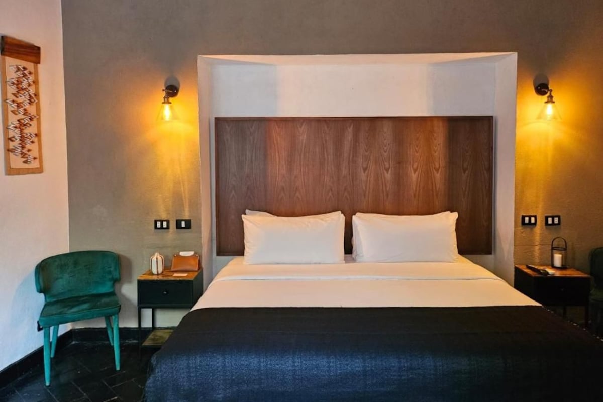 Quarto de hotel com uma cama grande, duas luminárias de cabeceira, uma mesinha de cabeceira, uma cadeira verde e um aplique na parede.