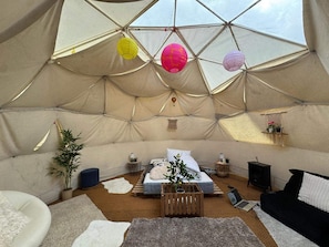 Acolhimento em tenda aconchegante, com cama, mesas de cabeceira, abajur, laptop e planta em vaso.