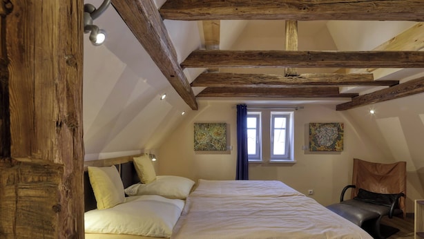 Quarto com teto inclinado, vigas de madeira aparentes, cama com almofadas, uma cadeira e obras de arte nas paredes.
