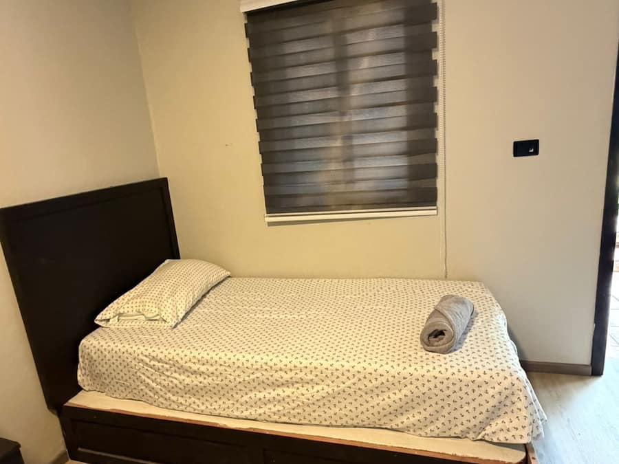 Uma cama de solteiro com cabeceira, uma janela com persianas e um interruptor de luz na parede.