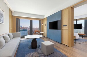 Quarto de hotel moderno com uma janela grande, um sofá, uma mesa redonda e uma televisão.