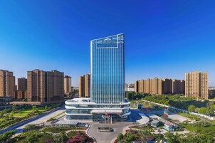 Um edifício moderno alto com fachada de vidro, cercado por área ajardinada com caminhos e vegetação.