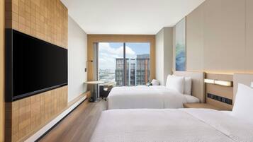 Quarto de hotel com duas camas, televisão de tela plana na parede, uma mesinha e vista para a paisagem urbana por meio de grandes janelas.