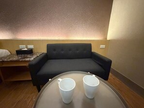 Quarto com sofá preto, uma mesa pequena com duas canecas brancas e uma mesinha de cabeceira de madeira com uma caixa de lenços.