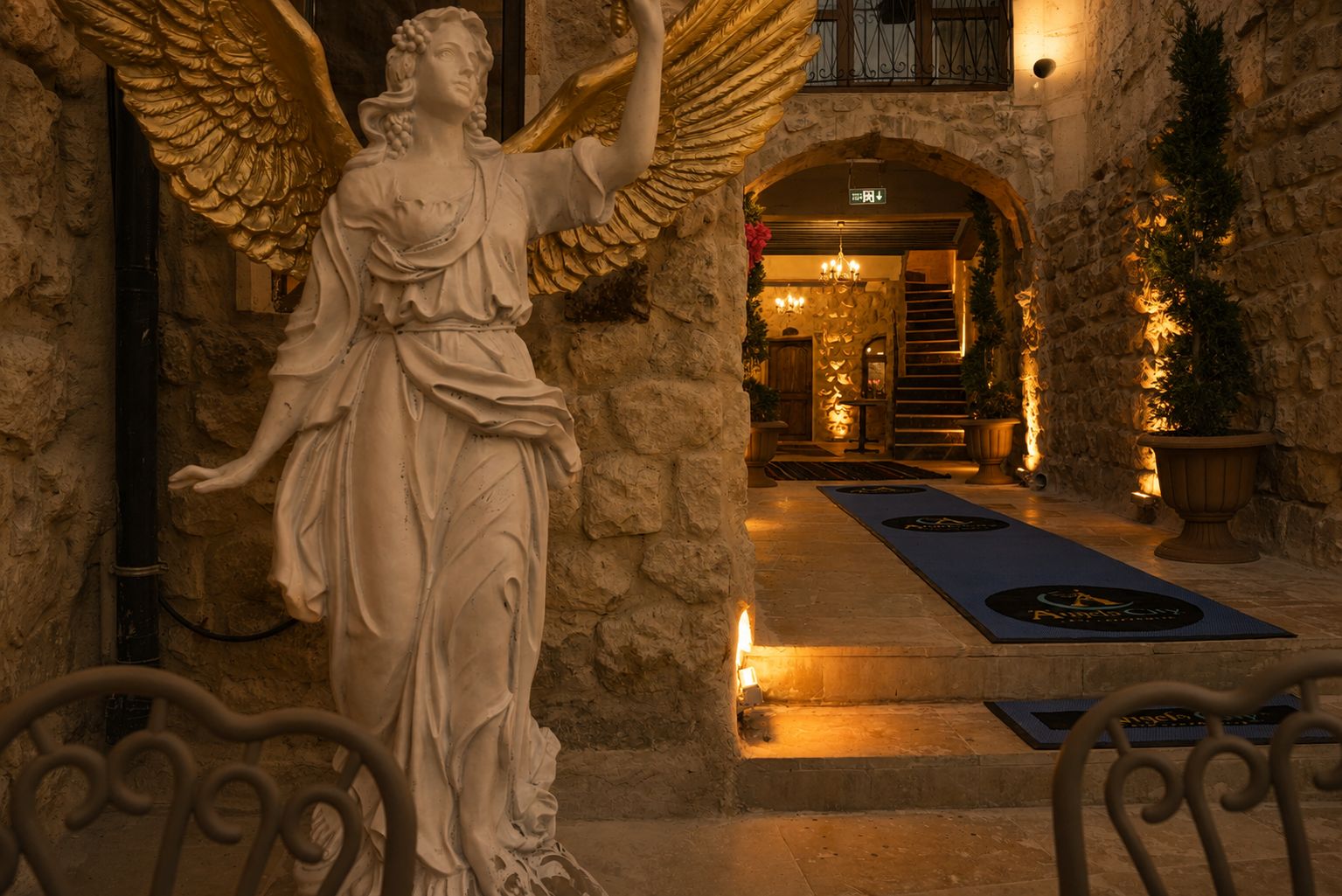 Uma estátua de anjo com asas abertas, uma parede de pedra, uma escadaria que leva ao interior e um tapete azul com um logotipo no chão.