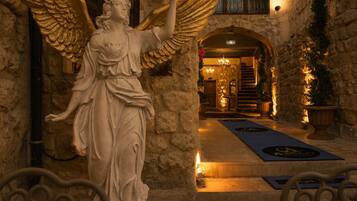 Uma estátua de anjo com asas abertas, uma parede de pedra, uma escadaria que leva ao interior e um tapete azul com um logotipo no chão.