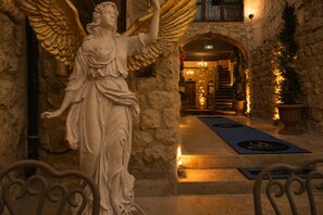 Uma estátua de anjo com asas abertas, uma parede de pedra, uma escadaria que leva ao interior e um tapete azul com um logotipo no chão.