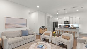 Uma sala de estar moderna com um sofá, poltronas e uma mesa de centro. Uma cozinha com armários e eletrodomésticos brancos é visível ao fundo.