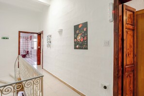 Um corredor com porta de madeira, sinal de saída de incêndio, luminária na parede e quadro decorativo com flores.
