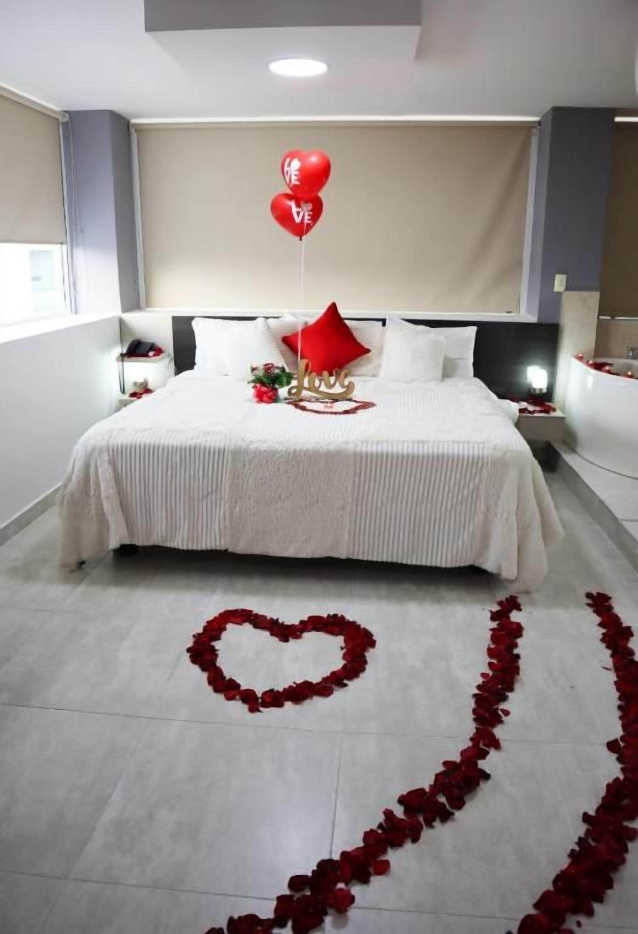 Quarto de hotel com uma cama grande, um arranjo de rosas em formato de coração no chão e um balão vermelho com a palavra 'LOVE'.