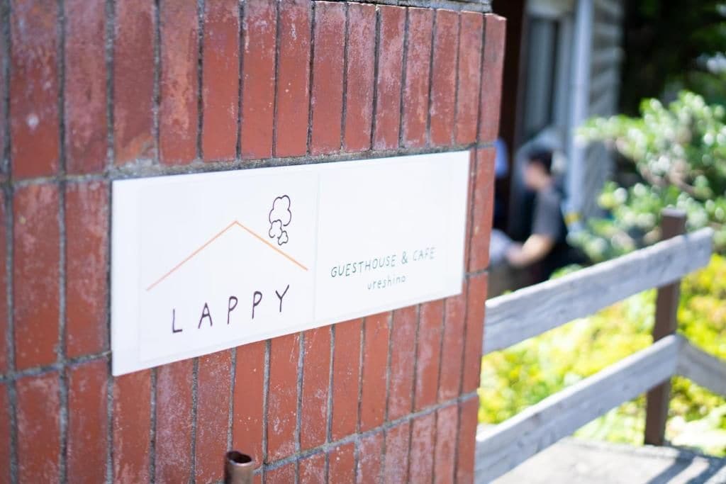 Parede de tijolos com uma placa que diz 'LAPPY GUESTHOUSE & CAFE' e um pequeno desenho de uma montanha.