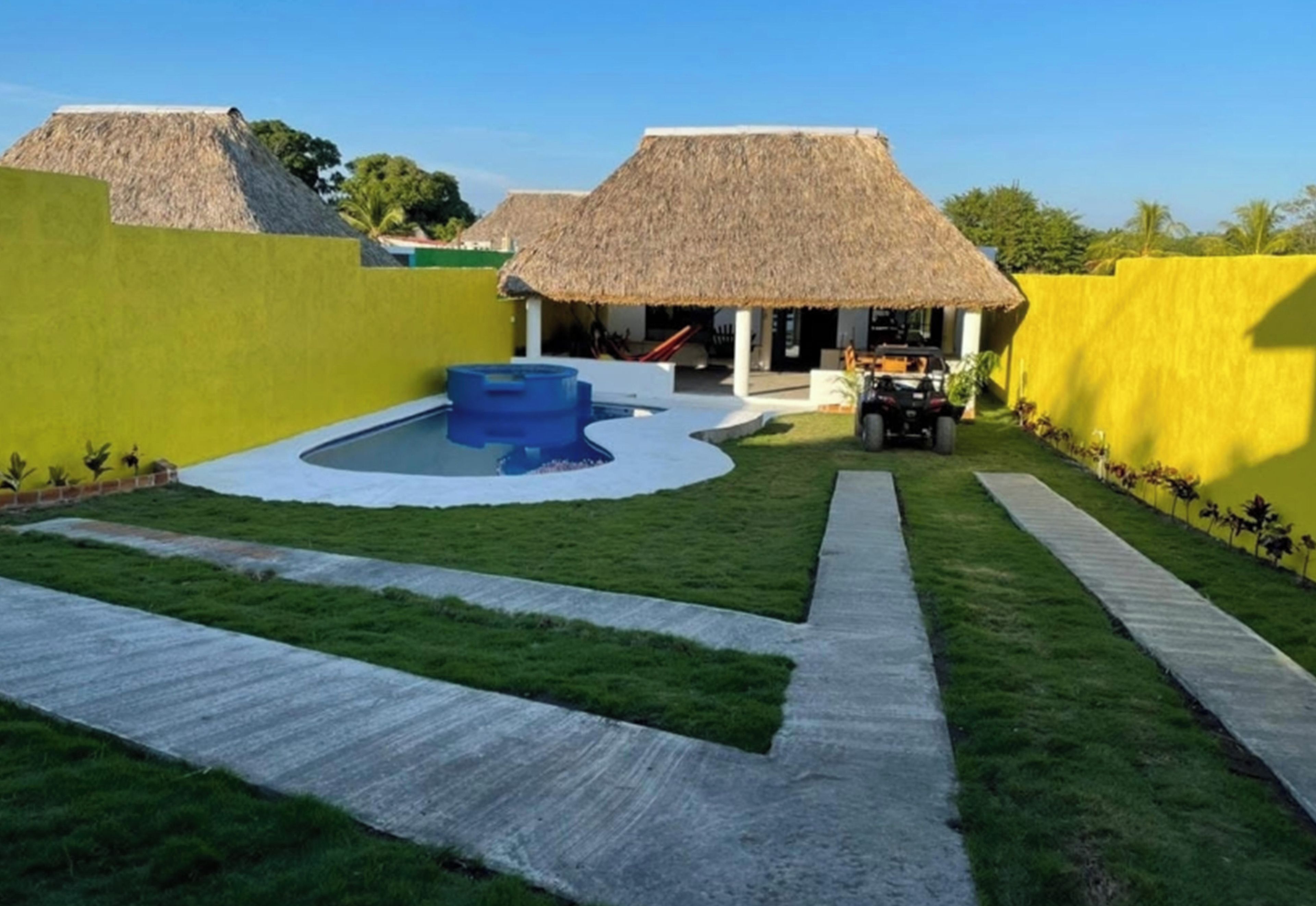 Uma área de piscina com uma banheira de hidromassagem azul, rodeada por uma estrutura com telhado de palha e uma parede amarela.