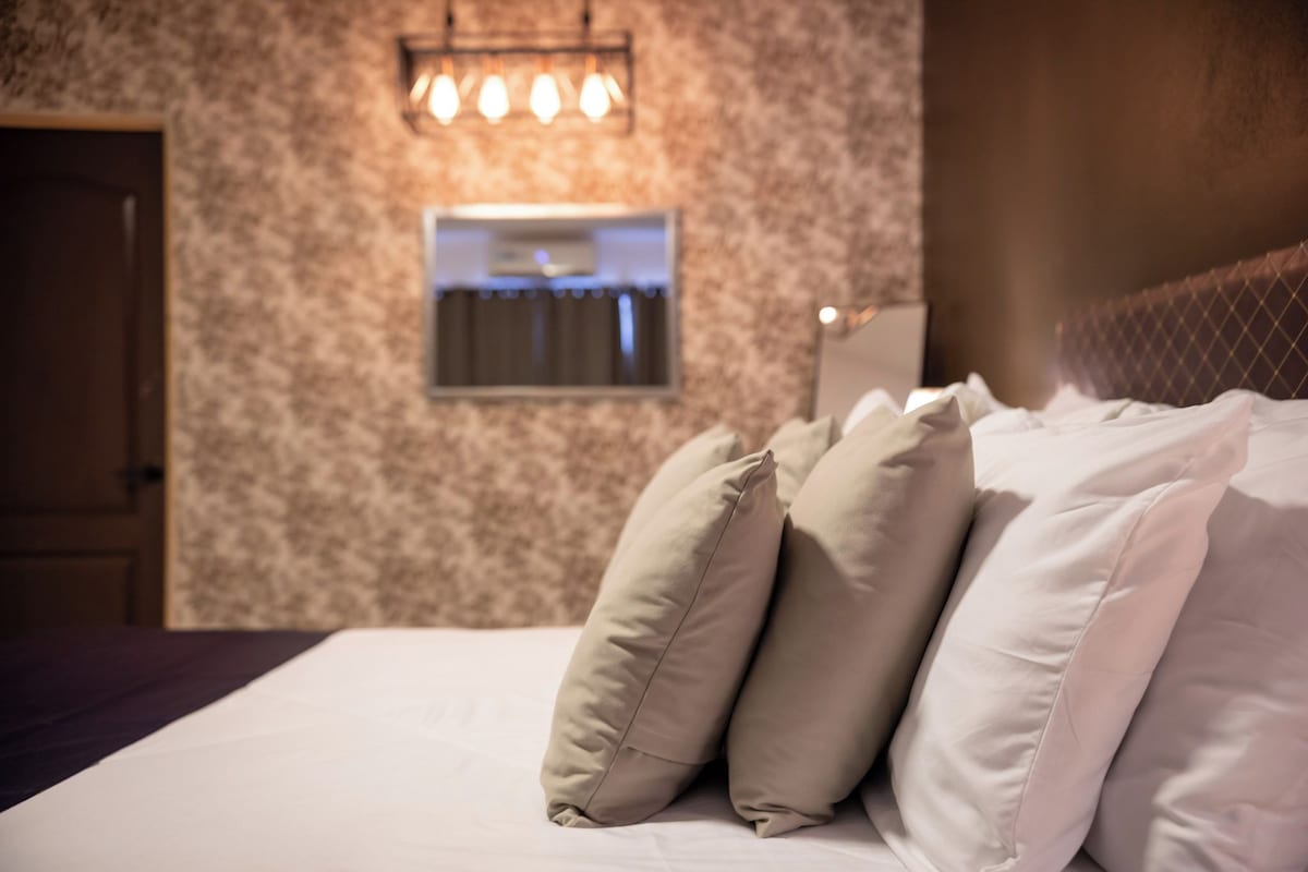 Quarto de hotel com cama, almofadas, mesa de cabeceira, abajur e luminária na parede.