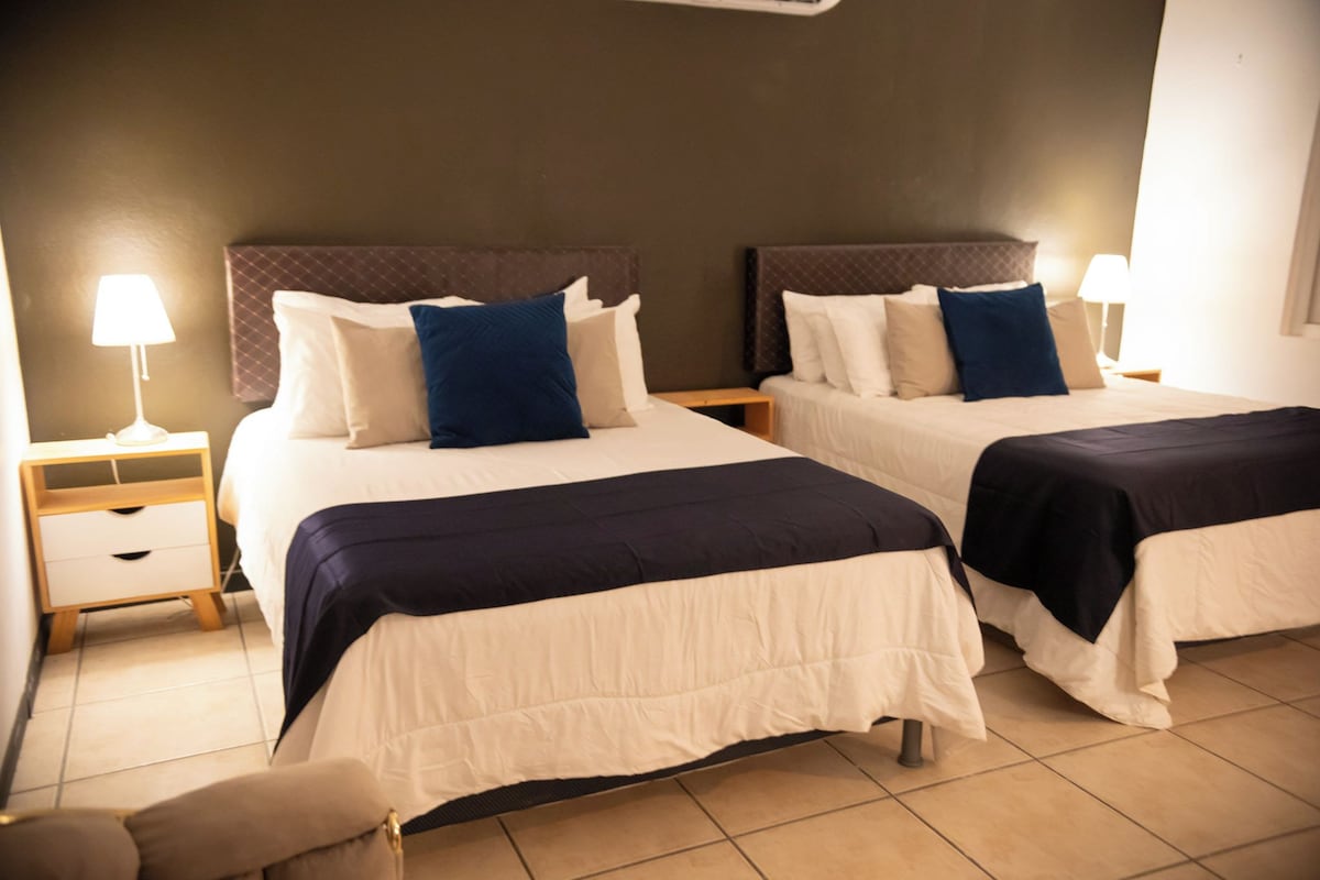 Quarto de hotel com duas camas, cada uma com cabeceira, mesas de cabeceira, luminárias e uma cadeira.