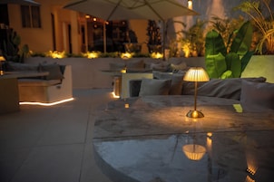 Uma área externa com assentos acolchoados, uma luminária de mesa e um grande guarda-sol.