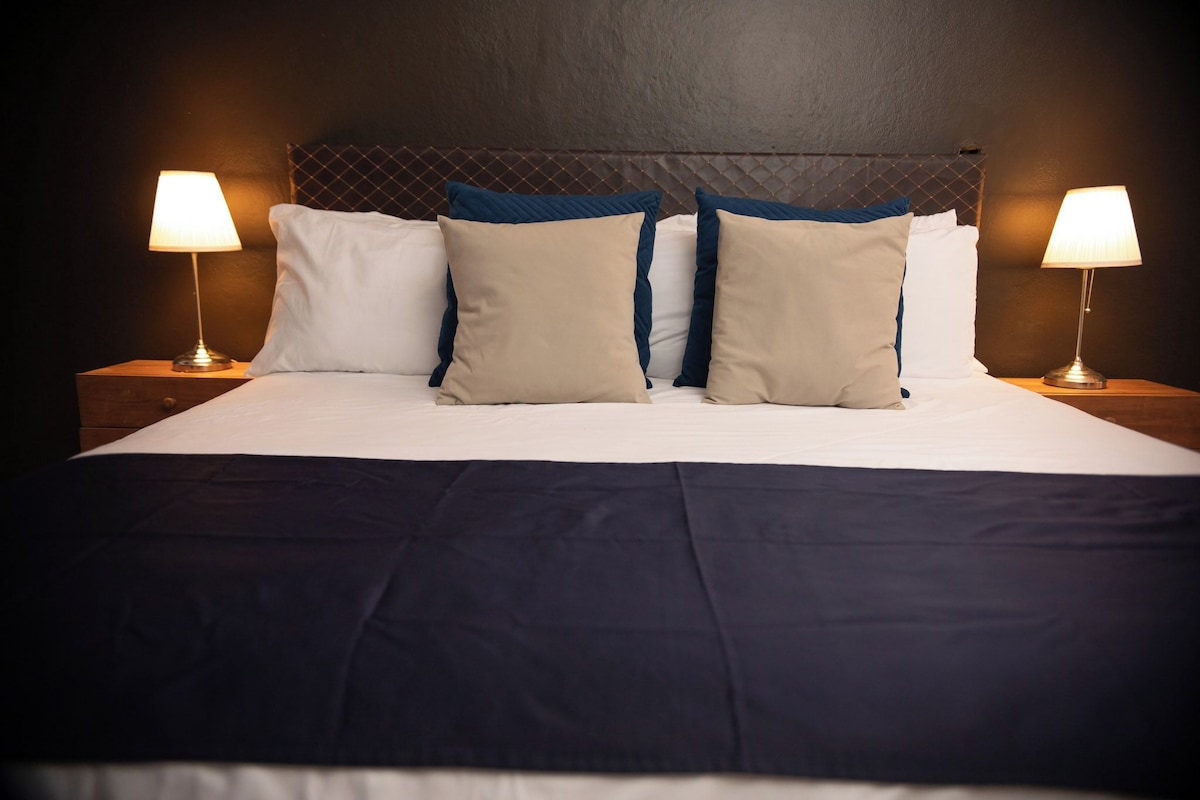 Cama bem arrumada com colcha azul escura, dois travesseiros brancos e dois travesseiros bege com detalhes azuis. Duas luminárias de cabeceira em mesas de cabeceira de madeira.