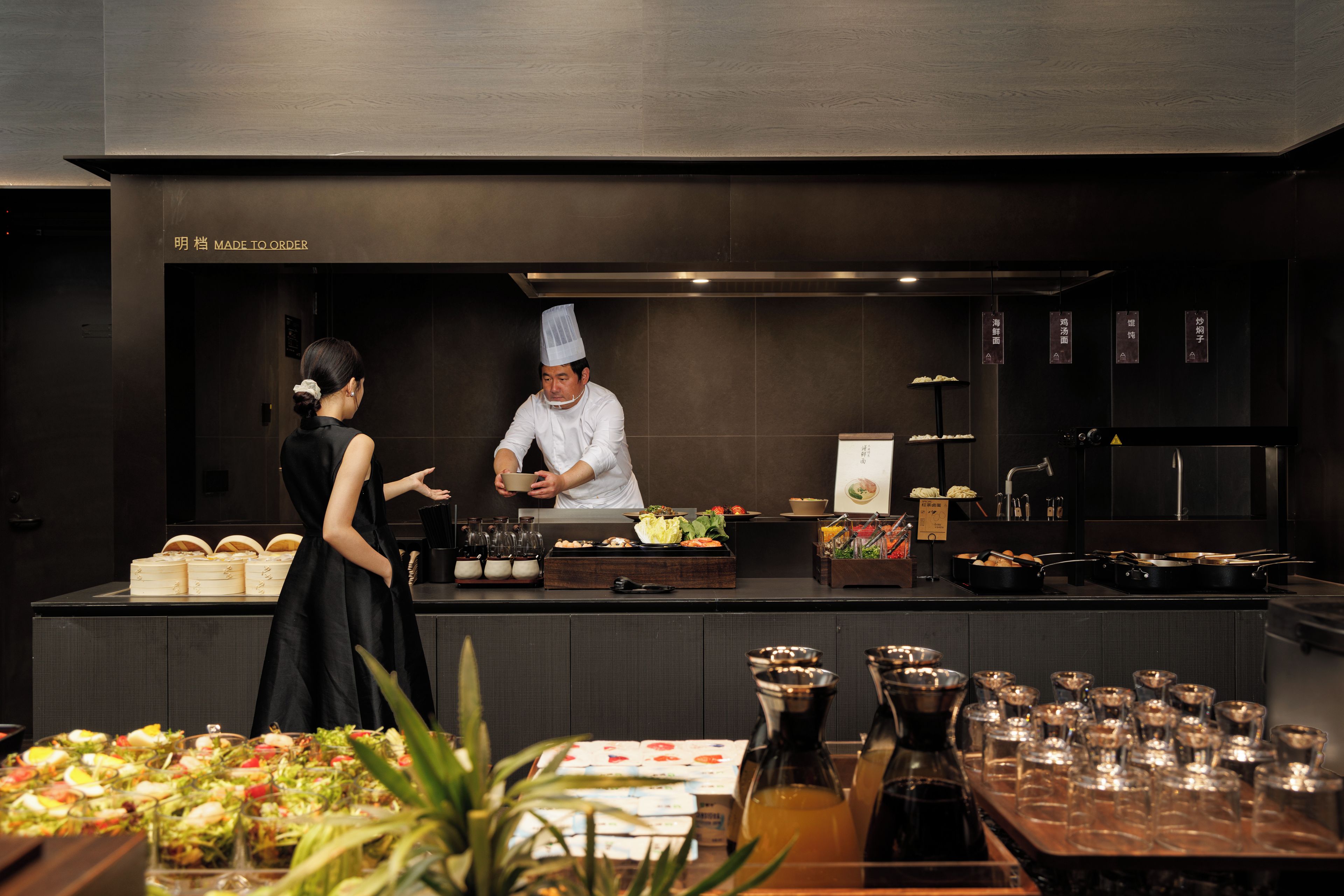 Um chef em cozinha profissional, uma mulher ao lado dele, mesa de buffet com diversos pratos e um quadro de menu ao fundo.